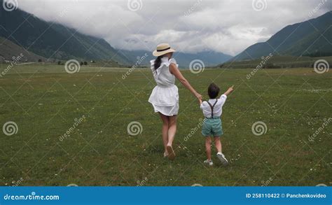 Мать женщина в белом платье бежит вместе с сыном через зелёное поле в горах летом есть