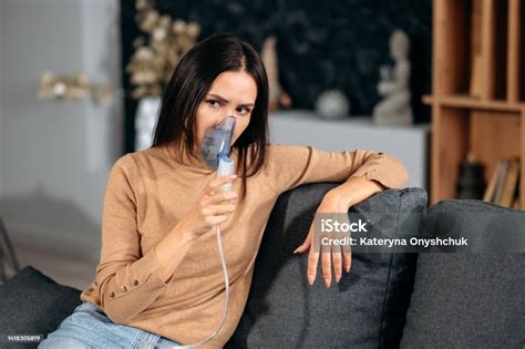 감기와 독감 개념 흡입기를 가진 Ill 백인 젊은 여성 집에서 흡입을하는 아픈 화가 난 소녀 그녀는 치료를 위해 분무기와 흡입기를 사용하여 격리 된 집에서 소파에 앉아