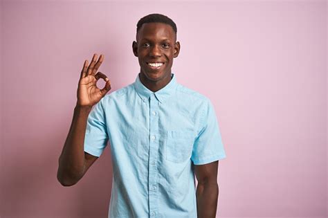 고립 된 분홍색 배경 위에 서있는 파란색 캐주얼 셔츠를 입은 아프리카 계 미국인 남성이 손과 손가락으로 확인 사인을하고 긍정적 인 미소를 짓고 있습니다 성공적인 식입니다
