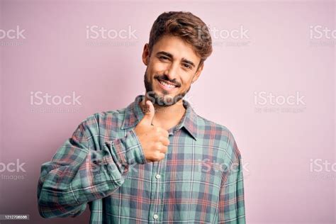 분홍색 배경 위에 서있는 캐주얼 셔츠를 입은 수염을 가진 젊은 잘 생긴 남자는 행복한 엄지 손가락으로 손을 제스처합니다 성공을 보여주는 카메라를 보고 표정을 승인합니다