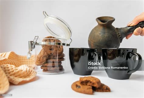 여성의 손은 흰색 배경에 검은 컵에 어두운 점토 Cezve에서 향기로운 커피를 부어 것입니다 초콜릿 칩 쿠키 의 항아리 이른 화창한 아침에 사랑이 가득한 아침 식사 가을에