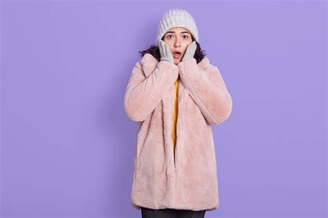 따뜻한 모피 코트와 모자를 쓰고 라일락 벽에 서서 뺨을 손으로 덮고 큰 눈으로 카메라를 바라보며 깜짝 놀랐습니다 20 24세에