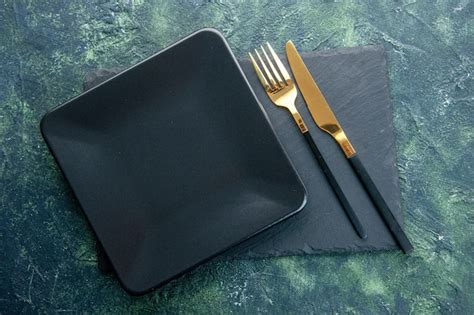 Вид сверху черная квадратная тарелка с золотой вилкой и ножом на темном ...