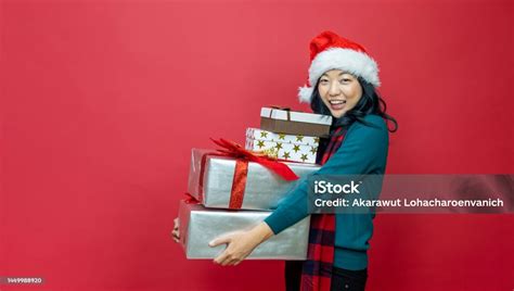 따뜻한 크리스마스 스웨터와 산타 모자를 쓴 예쁜 미소 짓는 아시아 여성이 계절 축하 개념을 위한 빨간색 배경으로 선물 상자를
