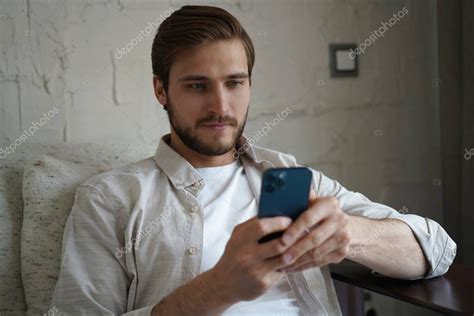 Красивый мужчина в обычной футболке улыбается и держит мобильный телефон в руках сидя на диване