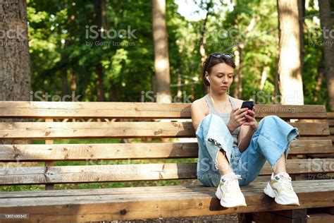 스마트 폰과 헤드폰으로 야외에서 벤치에 앉아있는 쾌활한 밀레니얼 금발 십대 소녀 공원에서 젊은 여학생 미소와 휴식 휴대용 통신 기기에 대한 스톡 사진 및 기타 이미지
