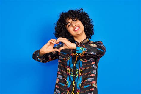 심장 기호와 손으로 모양을 보여주는 사랑에 미소 고립 된 파란색 배경에 화려한 셔츠를 입고 곱슬 머리를 가진 젊은 아랍 여성 로맨틱 한 개념 개념에 대한 스톡 사진 및 기타