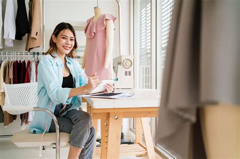 테일러링 아틀리에에서 옷을 작업하는 자신감 넘치는 아시아 재단사 여성의 초상화 젊고 매력적인 여성 패션 디자이너가 미소를 지으며 드레스 온 마네킹을 배경으로 카메라를