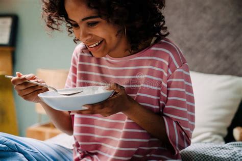 Menina Negra Rindo E Comendo Cereal Enquanto Sentada Na Cama Foto De Stock Imagem De