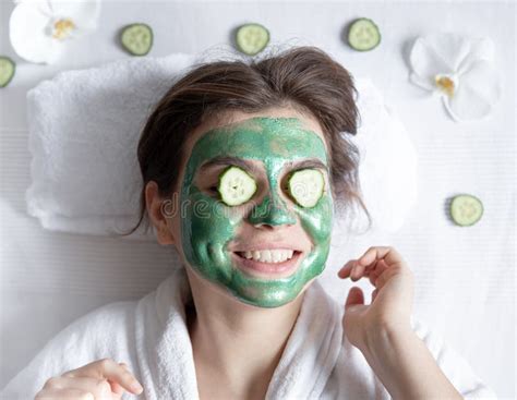 Funny Young Woman With A Cosmetic Mask On Her Face And Cucumbers On Her
