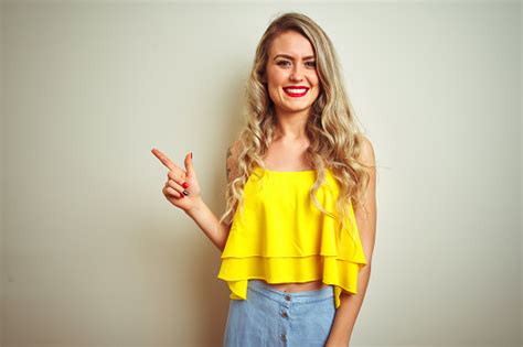 흰색 고립 된 배경에 서있는 노란색 티셔츠를 입은 젊은 아름다운 여성은 얼굴에 행복하고 자연스러운 표정으로 손과 손가락으로 가리키는 얼굴의 미소로 명랑합니다 T 셔츠에 대한