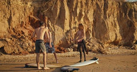 Three Bare Chested Guys In Shorts Stand On A Rocky Seashore On The Sand