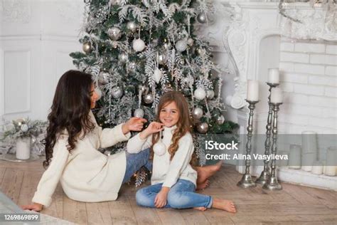 새해 전야에 행복한 가족 젊은 아름다운 어머니와 흰색 아늑한 스웨터와 담요에 작은 귀여운 딸은 장식 된 크리스마스 트리 아래에서 재미와 포옹을합니다 축제 홈 클래식 인테리어