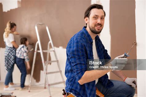 웃는 갈색 머리 남자가 작은 롤러로 벽을 갈색으로 칠합니다 그의 뒤에는 뒤에서 그의 아내와 딸이 있습니다 그들은 모두 수리에 관여합니다 35 39세에 대한 스톡 사진 및