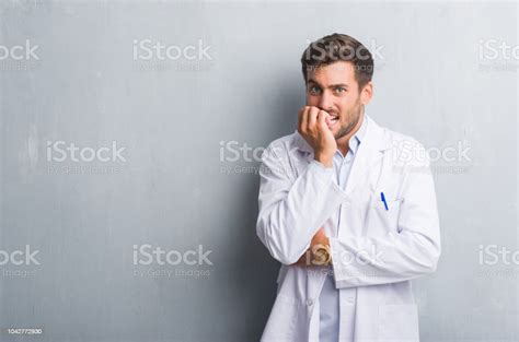 강조 하 고 손톱을 물어 뜯고 입에 손을 긴장을 찾고 흰색 코트를 입고 회색 그런 지 벽에 잘생긴 젊은 전문 남자 불안