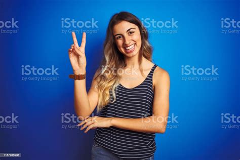파란색 고립 된 배경 위에 줄무늬 티셔츠를 입은 젊은 아름다운 여성은 승리의 표지판을하고있는 카메라에서 행복한 얼굴 윙크와 미소