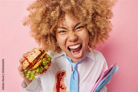 Outraged Curly Haired European Woman Feels Angry And Hungry Eats Appetizing Burger Holds Folders