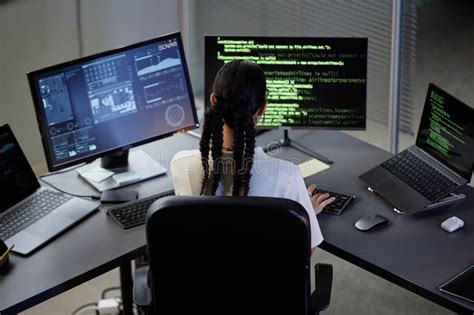 Back Of Female It Programmer Coding On Multiple Computers At Desk In