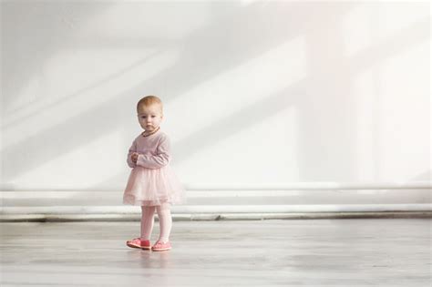 스웨터와 푹신한 치마를 입은 어린 아기가 회색 벽에 대고 커다란 빈 홀에 서서 카메라를 들여다봅니다 수평 형식입니다 텍스트 배치 12 17 개월에 대한 스톡 사진 및 기타