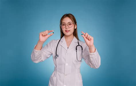 젊은 아시아 의사는 파란색 배경에 의료 유니폼 실험실 코트와 청진기를 착용합니다 두 손에 알약을 보여주는 미소 짓는 여성 의사 의료 약국 또는 건강 보험 개념 25 29세에