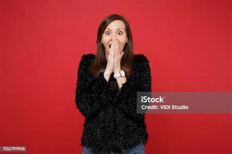 깜짝 놀란의 초상화는 스튜디오에서 밝은 빨간색 벽 배경에 고립 된 손으로 입을 덮고 검은 모피 스웨터에 젊은 여자 사람들은