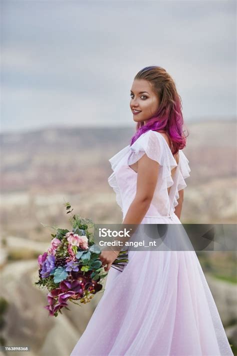 그녀의 손에 꽃의 아름다운 꽃다발을 가진 여자는 새벽 일몰의 광선에 산에 서있다 여자 몸에 아름 다운 흰색 긴 드레스입니다 핑크 머리와 완벽한 신부 결혼식에 대한 스톡 사진