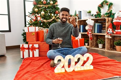 2022 년 새해를 축하하는 크리스마스 트리에 앉아 수염을 가진 젊은 히스패닉 남자는 분노와 손을 들고 미친 소리 분노와 좌절