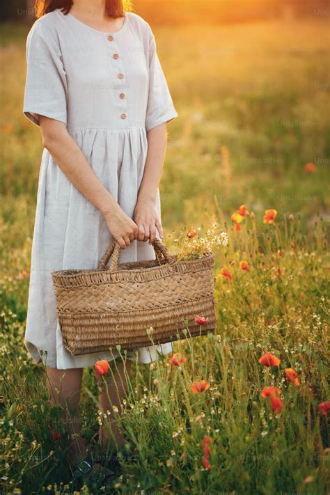 린넨 드레스를 입은 세련된 소녀는 일몰 빛 아래 초원에서 양귀비 꽃이 있는 소박한 짚 바구니를 들고 있습니다 여름 들판에서 야생화를 모으고 휴식을 취하는 보호 여성 대기