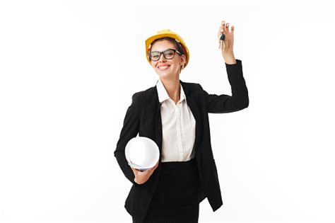 안경과 노란색 헬멧을 쓰고 미소 짓는 예쁜 사업가가 흰색 배경 위에 새로운 평면의 계획과 열쇠를 들고 카메라를 즐겁게 바라보고 있습니다 25 29세에 대한 스톡 사진 및