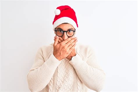 고립 된 배경 위에 크리스마스 모자와 안경을 쓰고 있는 젊은 잘생긴 남자는 실수로 손으로 입을 덮는 충격을 받았습니다 비밀 개념입니다 가리기에 대한 스톡 사진 및 기타
