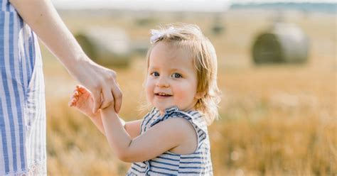 줄무늬 드레스를 입은 행복하고 즐거운 어린 소녀의 감동적인 초상화 일몰의 광선에 건초 더미가있는 밀밭에서 함께 걷는 동안 엄마의 손을 잡고 여름 유년 시대 사진