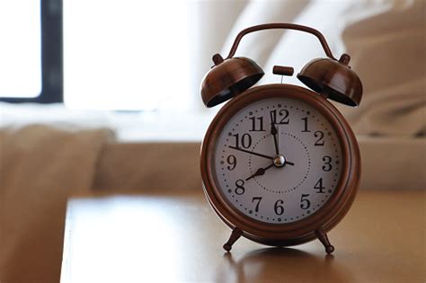 브론즈 메탈 케이스의 클로즈업 이미지 레트로 알람 시계 더블 벨 협탁에 흰색 다이얼이 있는 아날로그 디스플레이 시계 구겨진 침대 시트와 베개가 있는 더블 침대 전경에 초점