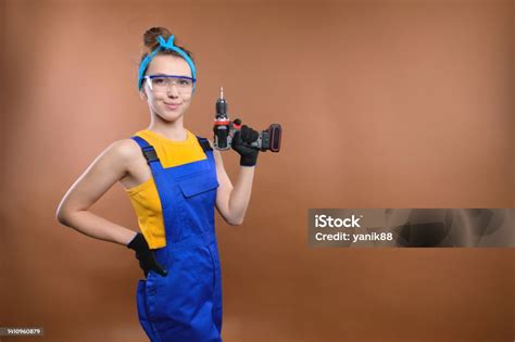 양성 평등 작업장에서 일하는 유니폼을 입고 전기 드릴 스크루 드라이버를 손에 들고 웃는 젊은 여성의 초상화 갈색 배경에 스튜디오