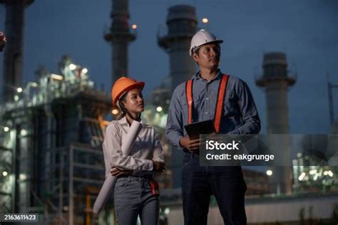 석유 화력 발전소 앞에 서서 청사진을 들고 밤에 일하는 아시아 팀 남성과 여성 엔지니어 노동자는 유니폼 헬멧 작업 타워 발전소