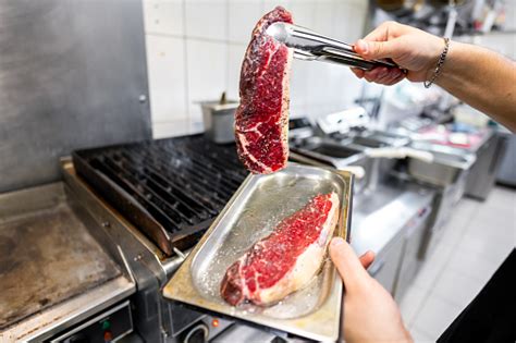 집게를 사용하여 마블링이 있는 생 스테이크를 전문 주방의 뜨거운 그릴에 올려놓는 셰프의 손 스테이크는 그릴에 닿으면서 지글지글 끓어 고기의 질감과 품질을 강조합니다 고기에
