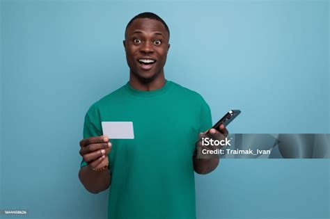 검은 피부를 가진 30세의 잘생긴 미국 남성은 스마트폰과 복사 공간이 있는 배경에서 쇼핑을 위한 플라스틱 카드를 들고 기본 티셔츠를 입고 있다 인사장에 대한 스톡 사진 및