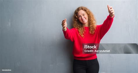 젊은 빨강 머리 여자 회색 그런 지 벽을 입고에 빨간색 스웨터 포옹에 대 한 두 팔을 벌려 웃 고는 카메라를 보고 명랑 식 수용