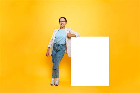 긍정적인 브라질 여성의 전체 길이 사진 프레젠테이션 발표 또는 광고를 위한 정사각형 흰색 모형 포스터 근처에 서 있고 엄지