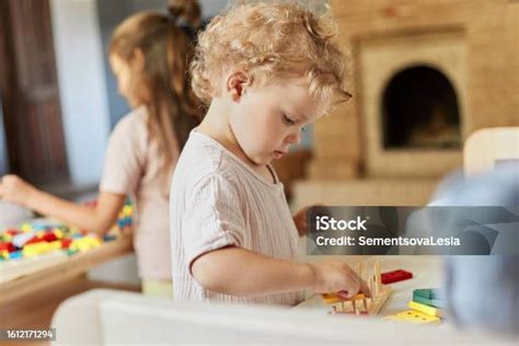 스마트 교육용 장난감 목조 건축물 건축 가사 보살핌과 학습 호기심 많은 물결 모양의 머리의 금발 유아 아기가 집 안에서 나무