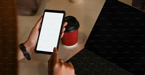 사무 장비로 둘러싸인 작업 책상 위에 흰색 빈 화면이 있는 잘린 검은색 스마트폰을 들고 있는 손의 자른 이미지 사진 Unsplash의 태국 이미지