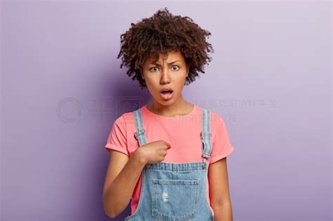 愤怒高清图片愤怒不满的黑皮肤女人的肖像指着自己，表情困惑，问为什么我被指控某件事，在室内穿 T 恤和工装裤模特有负面反应人像图片免费下载