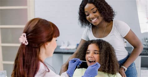 두려움없는 치과 치료 유니폼과 장갑을 낀 여성 치과 의사 어린 아프리카 계 미국인 소녀의 치아를 검사하는 동안 그녀의 어머니는 현대 소아 치과 진료소에서 그녀를 뒤에서