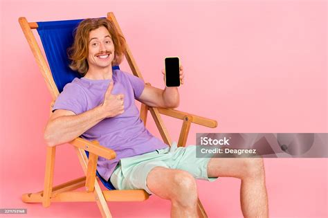 잘 생기고 유행 쾌활한 남자의 초상화는 의자에 앉아서 엄지 손가락 터치 스크린을 보여주는 장치를 들고 핑크 파스텔 색상 배경 위에 격리 30 34세에 대한 스톡 사진 및