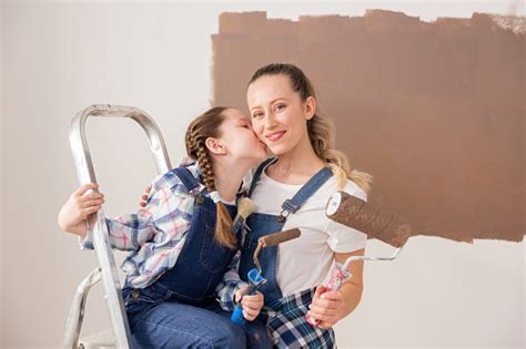 데님 작업복과 체크 무늬 셔츠를 입은 매우 귀여운 8 세 소녀가 사랑스러운 엄마에게 키스합니다 그녀의 가족이 아파트에서 개조를 시작했기 때문에 아이는 사다리에 앉아 있습니다