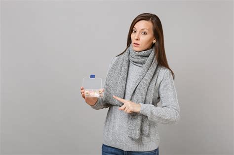 회색 스웨터 회색 벽 배경에 고립 된 매일 약 상자에 스카프 포인팅 인덱스 손가락에 젊은 여자 건강 한 라이프 스타일 아픈 질병 치료 추운 계절 개념입니다 복사 공간을 모의