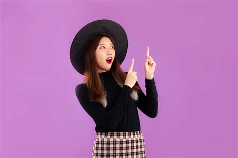 할로윈 테마 검은 긴 소매 티셔츠를 입은 젊은 아시아 여성이 마녀 모자를 쓰고 보라색 배경에 손가락 포이니팅을 가하고 있습니다