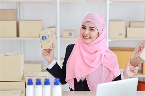 파란색 무슬림 정장을 입은 활동적인 아시아 여성이 앉아서 컴퓨터로 일하고 행복한 웃는 얼굴로 온라인 소포 상자 배달을 보여주고