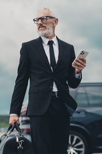 정장을 입은 성숙한 사업가가 도시 주차장에서 차 근처에서 전화와 가방을 들고 있습니다 50 54세에 대한 스톡 사진 및 기타 이미지 50 54세 Brand Name