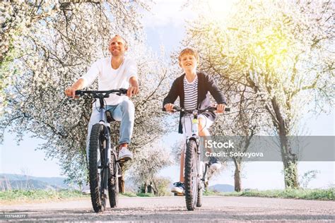 아버지와 아들은 꽃 나무 아래에 있는 시골 길에서 자전거를 타는 것을 즐겁게 합니다 건강 한 스포티 라이프 스타일 컨셉 이미지 2명에 대한 스톡 사진 및 기타 이미지
