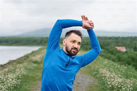 야외에서 운동하는 중년 스포츠맨 팔 근육을 따뜻하게 여름 우울한 날 경치가 좋은 도로에 서서 워밍업 스트레칭 파란색 재킷을 입고 수염이 있습니다 사진